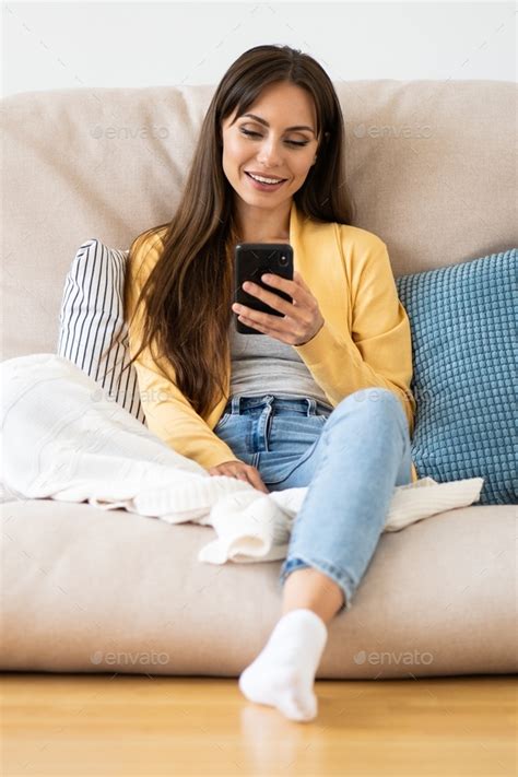 Smiling Millennial Woman Resting Sit On Sofa At Home Using Cellphone