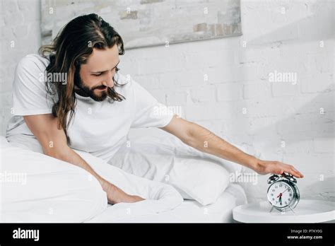 Cheerful Young Long Haired Man Turning Off Alarm Clock In Bedroom At