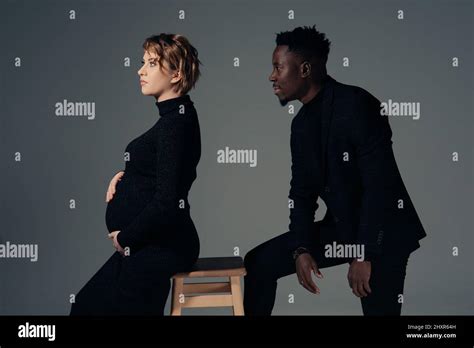 Young African Man And His Caucasian Pregnant Woman Sit Against Grey