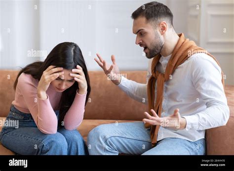 Aggressive Husband Shouting At Unhappy Wife Sitting On Sofa Indoor