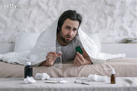 Unhappy Millennial Caucasian Guy With Stubble Lies On Bed Under Blanket