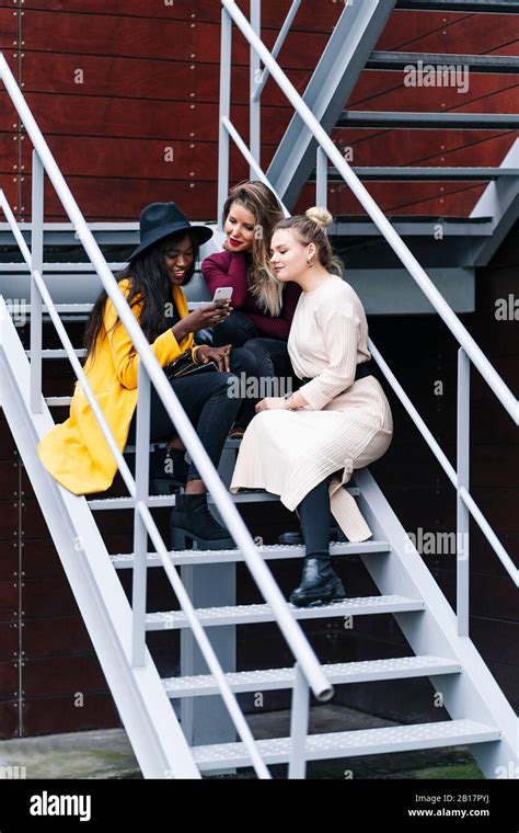 Girlfriends Sitting On Stairs Outdoors Looking At Mobile Phone Stock