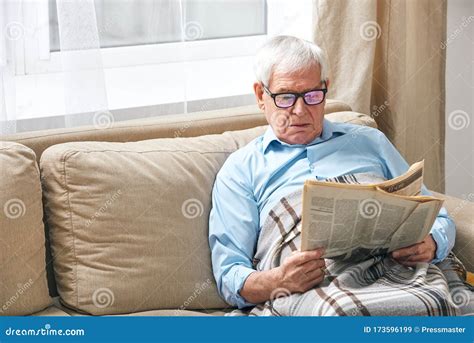 Senior Man Wrapped In Plaid Reading Newspaper While Relaxing On Couch