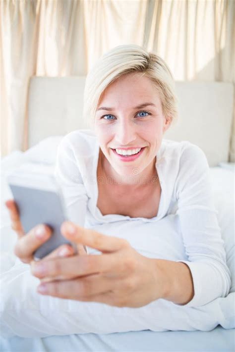 Smiling Blonde Woman Lying On The Bed And Texting With Her Mobile Phone