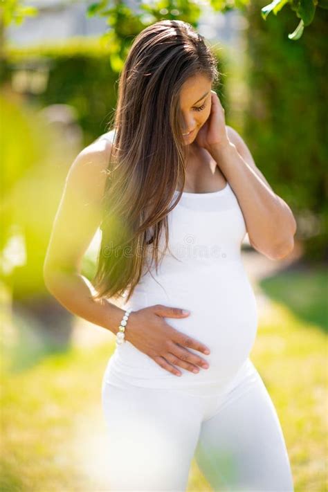 Happy Pregnant Woman Standing In Garden Holding Hands On Belly Stock