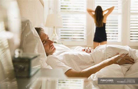 Wife Stretching At Morning Window Behind Husband Sleeping In Bed — Eyes