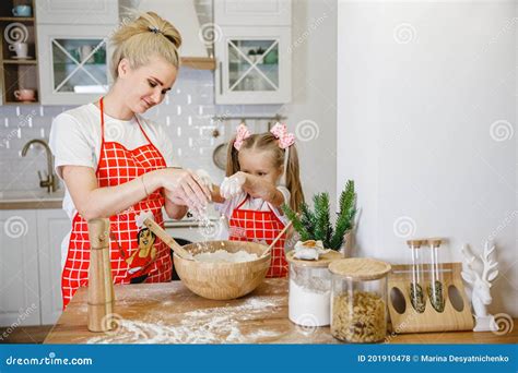 Blonde Mom And Daughter In Red Checkered Aprons Knead Dough In The