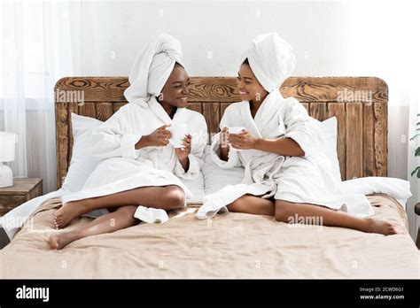 African American Ladies In Bathrobes Sitting On Bed Drinking Coffee