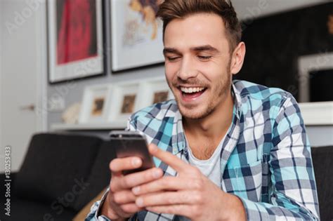 Young Laughing Man Sitting In Home And Looking At Phone Stockfotos