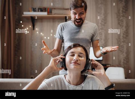 Angry Husband Shouting At Wife Sitting On Sofa And Covering Ears With
