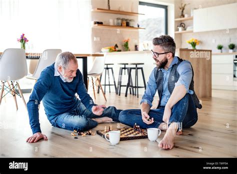 An Adult Hipster Son And Senior Father Sitting On Floor Indoors At Home