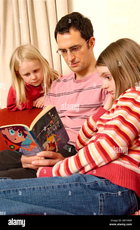 Father Reading Book To His Daughters On Sofa In Living Room At Home Uk