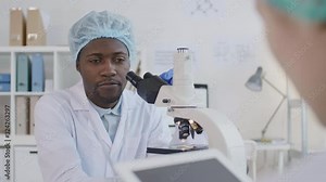 Back view of African man wearing special uniform sitting at desktop, using microscope and talking to his unrecognizable coworker sitting nearby and holding tablet