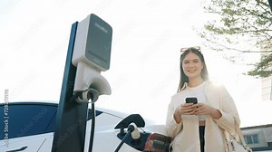 Slow motion young woman check battery status on smartphone while recharging EV car battery from charging station at city mall parking lot. Modern woman go shopping by electric vehicle. Expedient