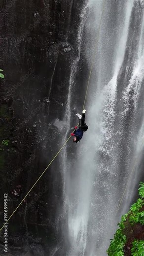 Cinematic drone view of a professional climber rappelling down a giant rushing waterfall. Epic aerial footage of extreme canyoneering adventure in a deep rainforest canyon with heavy mist