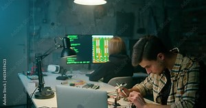 Man is Soldering in Electronics Workshop. Hacking Crime, Cyber War or IT Startup Concept. Young Coders Work on Technology Project in Dark Garage, Basement or Loft Office. 4K Wide Pan Right shot
