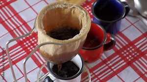 Coffee being filtered through a cloth strainer in enameled cups and homemade bread in the background on a checkered tablecloth. Making coffee with an cloth strainer also known as Mariquinha in Brazil