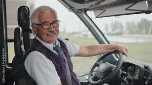 Medium portrait of cheerful senior bus driver in uniform sitting at driver seat with hands on steering wheel smiling to camera