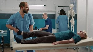 Elder person raising legs to stretch muscles in osteopathy cabinet, receiving support from male physiotherapist. Medic helping with stretching procedure exercise at rehabilitative physiotherapy.