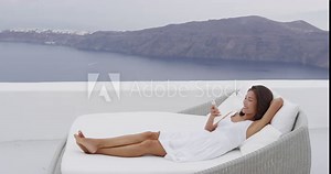 Elegant stylish woman taking selfie and texting using cell phone app relaxing in sofa on luxury terrace. Asian woman using smartphone on daybed on outdoor with Oia Santorini background enjoying summer