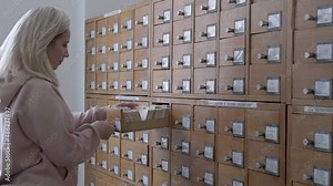 4K. A young caucasian blonde woman is looking for information in a file cabinet. Female student looking for necessary literature in library reference card catalog. Search documents in the card index