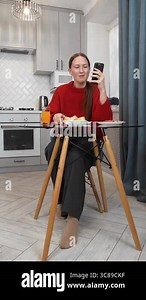 Young woman sitting at glass table in her modern kitchen enjoying breakfast while working with her smartphone, creating a cozy and connected morning routine. vertical video Stock Video Footage - Alamy