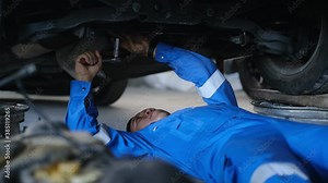Mechanic Asian man underneath car fixing repairing front axle beam automobile vehicle parts examining using tools equipment working hard in workshop garage support and service in overall work uniform