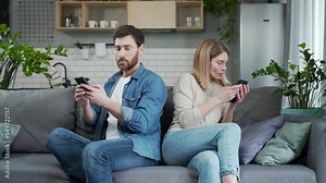 Married young couple sitting on couch back to back typing scrolling chatting in social network on smartphones at home Wife and husband ignoring checking each other screen Concept of trust in marriage