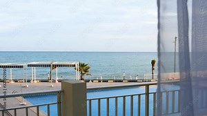Footage with handheld effect from the point of view of a person standing by the window looking at the blue sea or ocean with yacht through fluttering transparent white curtain on sunny summer day