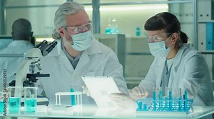 Mature Caucasian scientist in white coat, protective mask and gloves using microscope and talking with young female colleague as she working with digital tablet in laboratory