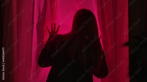 Back view of a curious woman standing near a window in a dark room looking out into the street, close up. Woman looking at flashing lights of police or other emergency service.