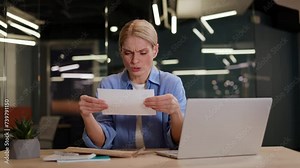 Woman opening letter envelope and getting disappointed while sitting at workplace with laptop in modern office. Blond female receiving notification from boss and learning that she got fired from work.
