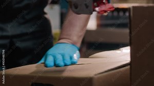 Cropped closeup shot of unrecognizable industrial male worker wearing blue nitrile gloves packaging cardboard boxes with manual scotch tape dispenser