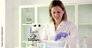 Positive scientist checks glass test tubes standing near electronic microscope. Woman smiles preparing for research in laboratory slow motion