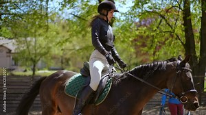 Tracking shot of graceful horse running in circle in paddock with equestrian exercising and assistant holding bridle. Wide shot confident Caucasian woman and purebred stallion training outdoors