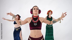 Portrait of confident talented women moving chest and hands in slow motion performing Middle Eastern dance at white background. Front view three Caucasian performers dancing belly dance
