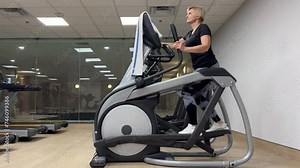 Middle-aged woman goes on an elliptical exercise machine. Happy 55-60 year old woman doing cardio workout. Active healthy lifestyle.