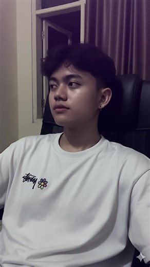 Buat gambar di atas tanpa merubah wajah dan rambutnya menjadi Potret jarak dekat seorang pria muda (berdasarkan foto referensi) duduk di kursi hitam di dalam ruangan pada malam hari, suasana yang tenang dan intim. Dia duduk dengan nyaman, punggung bersandar ringan di kursi, bahu rileks. Dia menatap ke depan kamera , ekspresi netral dengan tatapan percaya diri yang lembut , maskulinitas halus bercampur dengan kelembutan yang tenang. Kepala sedikit miring, menekankan garis rahang yang jelas Rambut