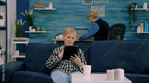 Focused senior grandmother relaxing sitting on sofa typing tablet browsing, searching informations, shopping online, while adult man working at laptop in background. Person using notepad gadget