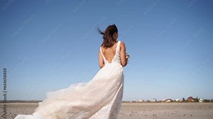 Pretty woman in gorgeous wedding dress walking at desert landscape, windy weather. Elegance bride with bouquet of fresh flowers waiting for a groom after wedding ceremony. Attractive model going.