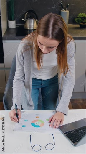 Top view young female employee working on project on laptop, standing at shared table in modern office kitchen. Business woman holding documents, involved in financial paperwork using computer e