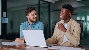 Two busy diverse male employees using laptop computer talking in office. Professional young business men workers team having conversation working on project sharing ideas sitting at work desk.