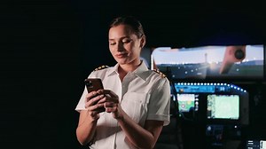 Front view of female pilot, stewardess standing in cabin, wearing uniform. Young, pretty woman looking at camera, smiling, holding,, using smartphone. Concept of aviation.