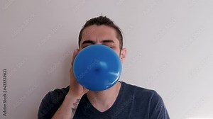 Inflate the blue balloon with mouth and then let it fly away. Young, cute Caucasian European man with beard and ponytail inflates balloon on white studio background.