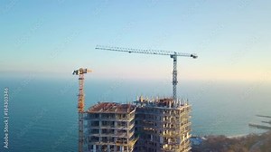 Aerial city view. Construction of a high-rise skyscraper on the ocean by two cranes. Even slow fly. The camera focusses the view on the high-rise crane and turns down.