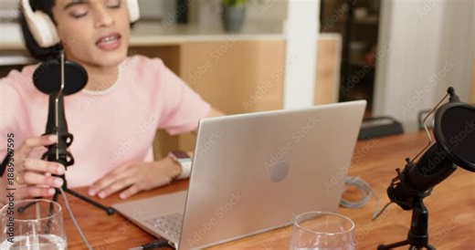 Man adjusting laptop and pop-filter mic, donning headphones, recording podcast at kitchen table