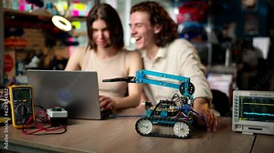 Two young happy engineers fixing a mechanical robot car in the workshop, computer programming