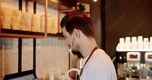 Young man using a point of sale system in a coffee shop. Male barista wearing face mask using till. Waiter registering client's order in a computer at coffee shop. Cashier at a coffee shop.