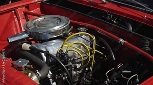 Clean and shiny old carbureted engine in engine compartment of red old car. Under sunlight is demo retro car without hood. Passing people are reflected in chrome air intake.
