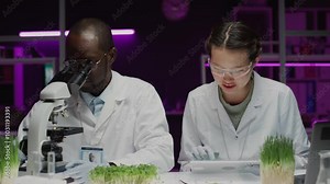 African American male botanist talking with Asian female colleague, then looking through microscope as she using digital tablet during plant genetics research in laboratory